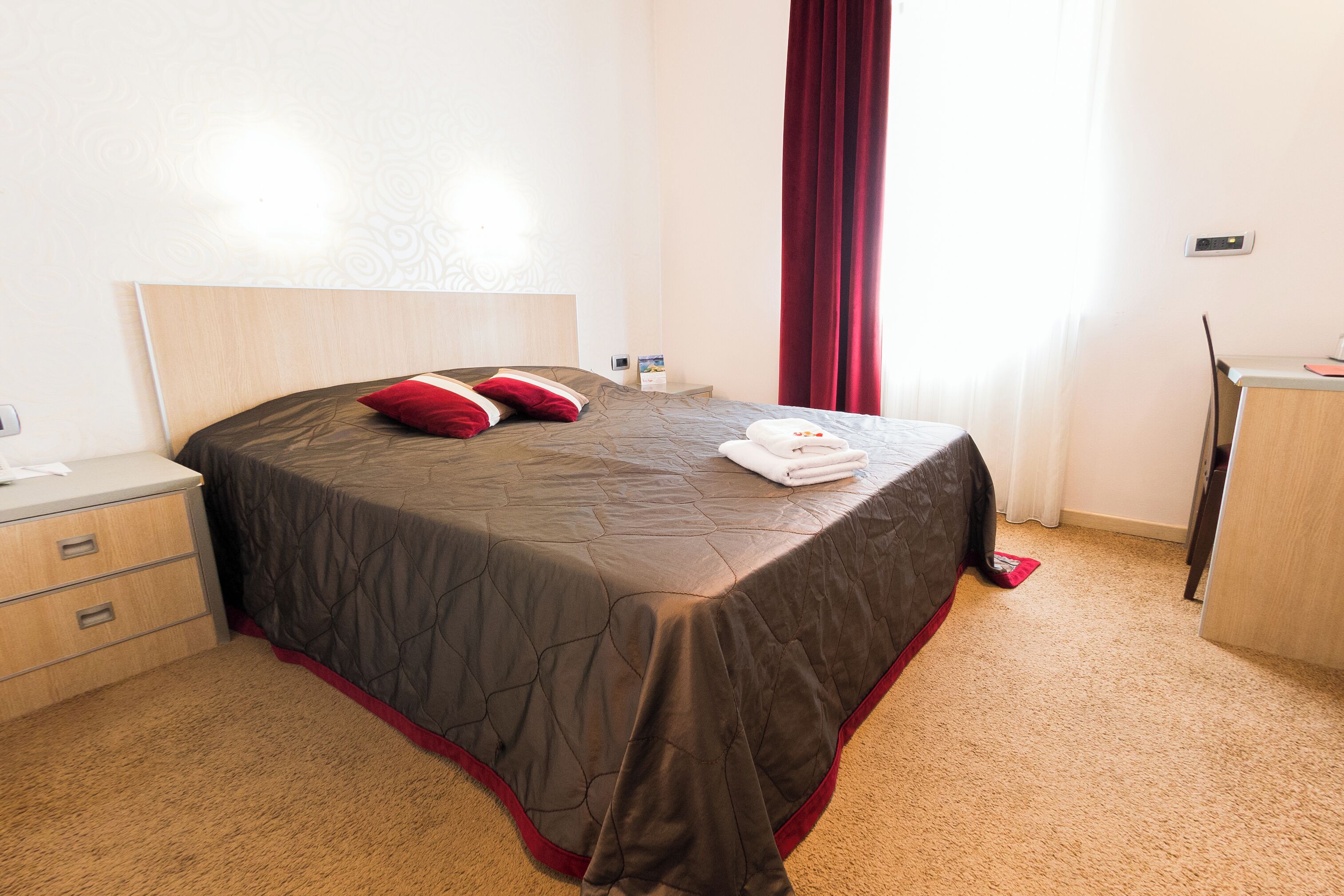 Cama bem arrumada com um edredom marrom, duas almofadas vermelhas e uma toalha branca em uma cabeceira de madeira.