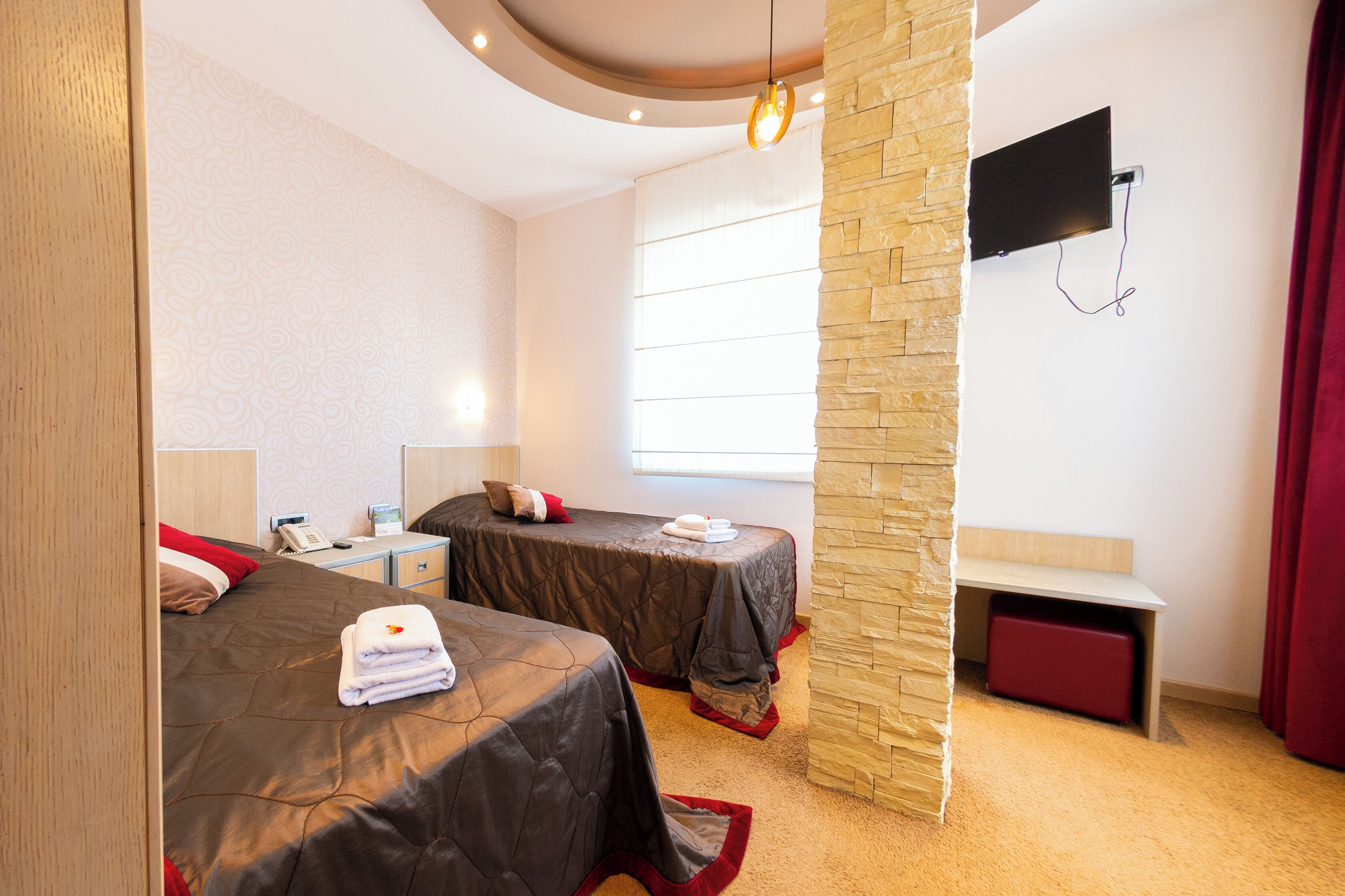 Quarto de hotel com duas camas, uma televisão fixada na parede, um pilar de pedra e um banco.