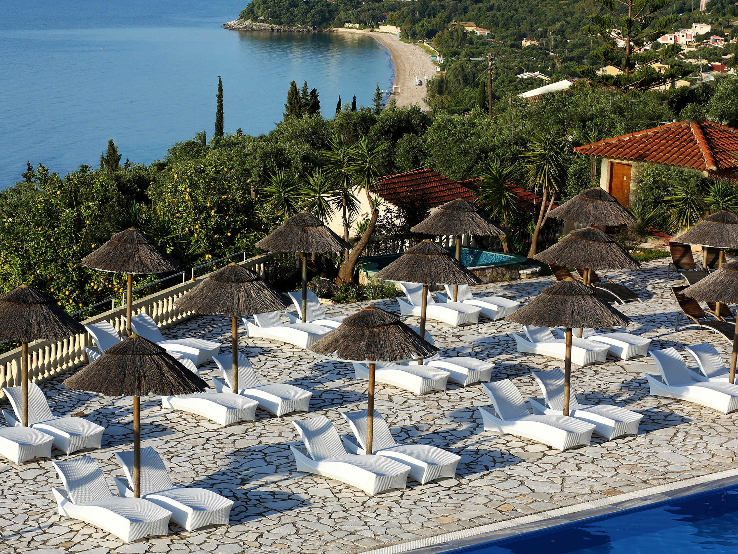 Área de piscina com cadeiras de banho brancas e guarda-sóis de palha, com vista para uma praia e uma paisagem montanhosa.