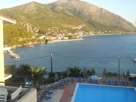 Uma área de piscina com espreguiçadeiras azuis e guarda-sóis, com vista para uma vila costeira e uma montanha.
