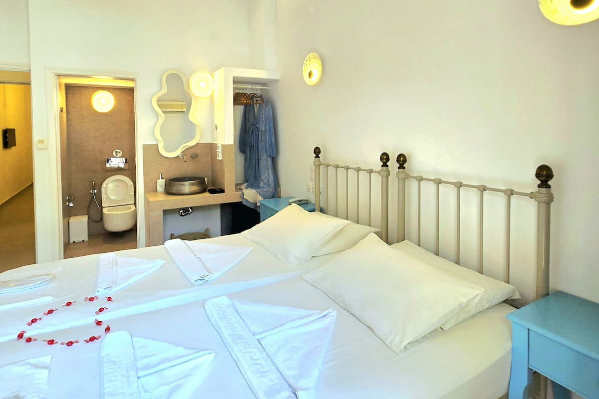 Quarto de hotel com uma cama, uma mesa de cabeceira, banheiro com vaso sanitário e pia, e um espelho.