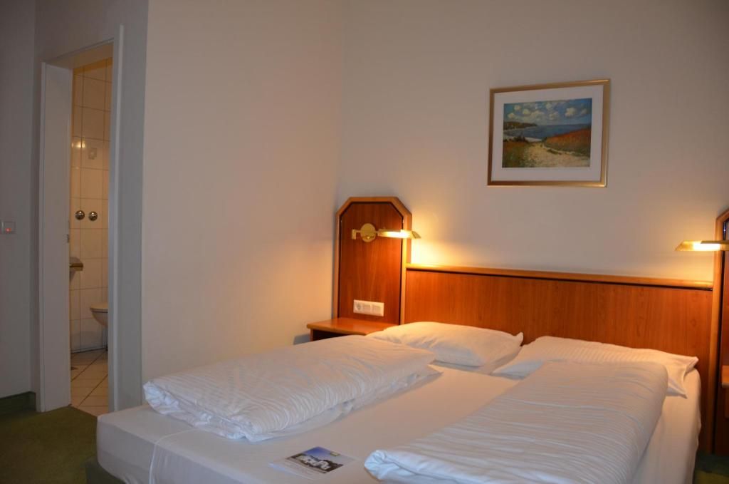 Quarto de hotel com cama de casal, cabeceira de madeira, abajures, quadro emoldurado na parede e porta de acesso a um banheiro.
