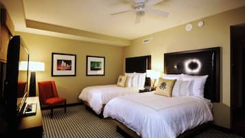 Quarto de hotel com duas camas, um ventilador de teto, uma cadeira vermelha, uma escrivaninha com abajur e quadros nas paredes.