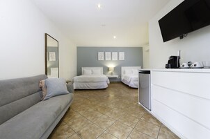 Quarto de hotel com sofá cinzento, duas camas de solteiro, cómoda branca, televisão na parede e máquina de café.
