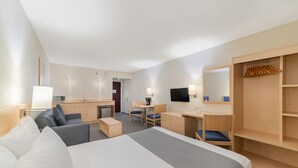 Quarto de hotel com cama, escrivaninha, poltrona, sofá, televisão, guarda-roupa e uma pequena cozinha.