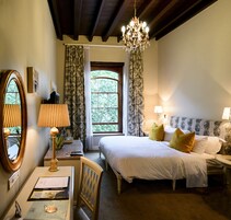 Quarto com cama, mesa de cabeceira, abajur, escrivaninha, cadeira e uma janela grande com cortinas.