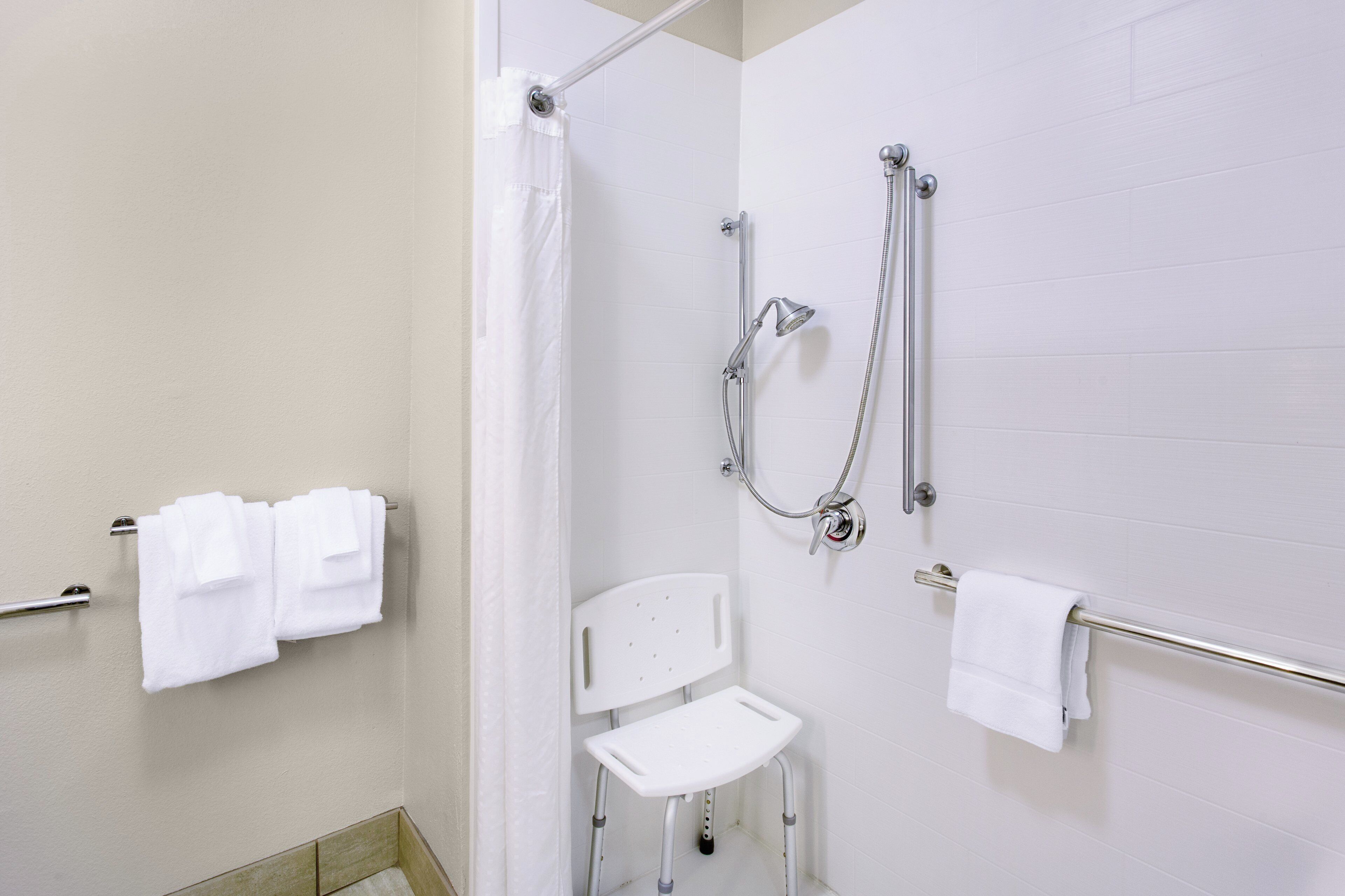 Casa de banho com chuveiro, uma cadeira branca e várias toalhas em porta-toalhas.