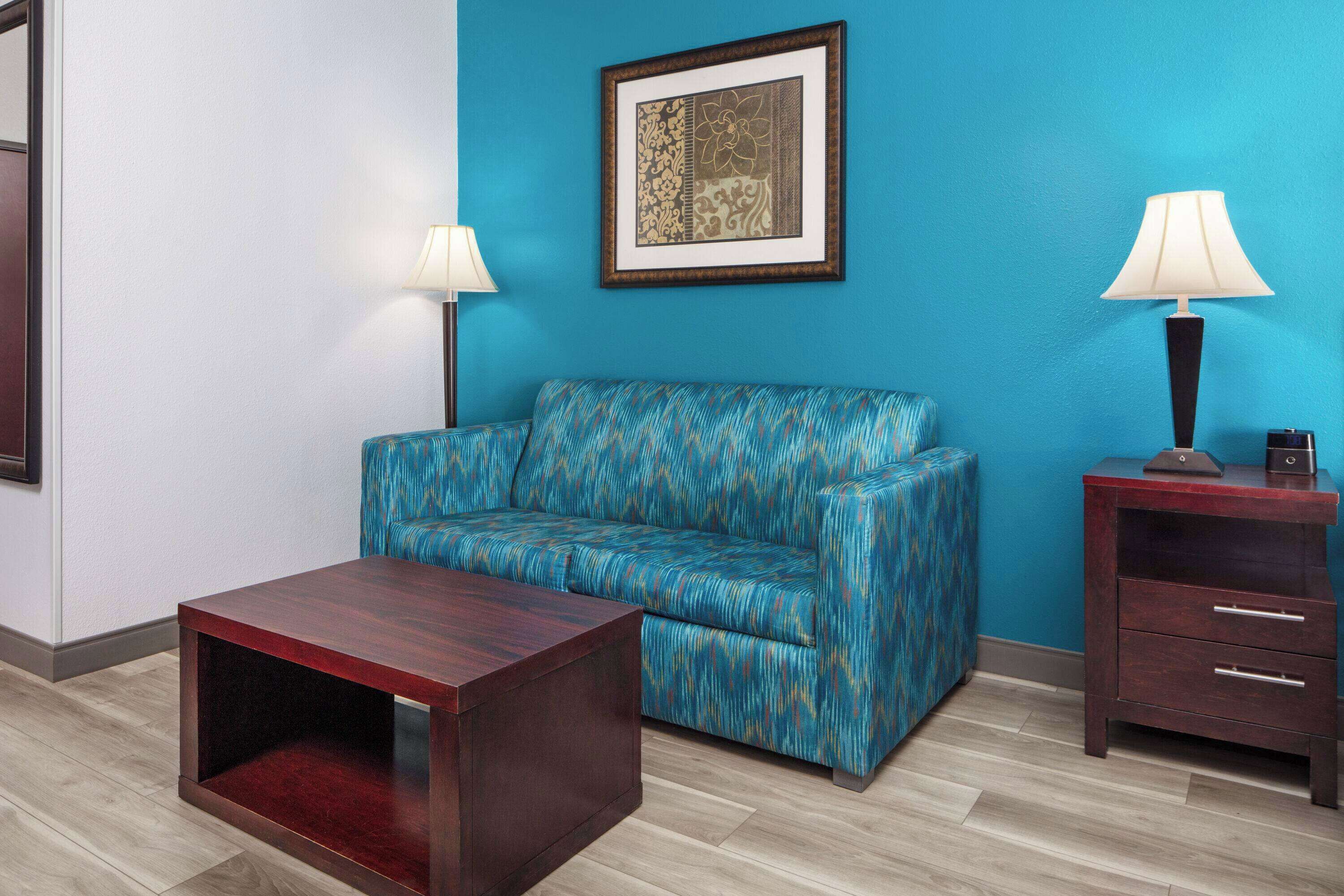 Um quarto com parede azul, sofá estampado, mesa de centro de madeira, mesa de cabeceira com abajur e quadro pendurado na parede.