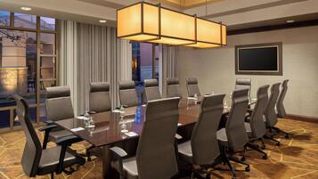 Uma sala de conferências com uma grande mesa, várias cadeiras, um lustre moderno, uma janela ampla com cortinas e uma tela na parede.