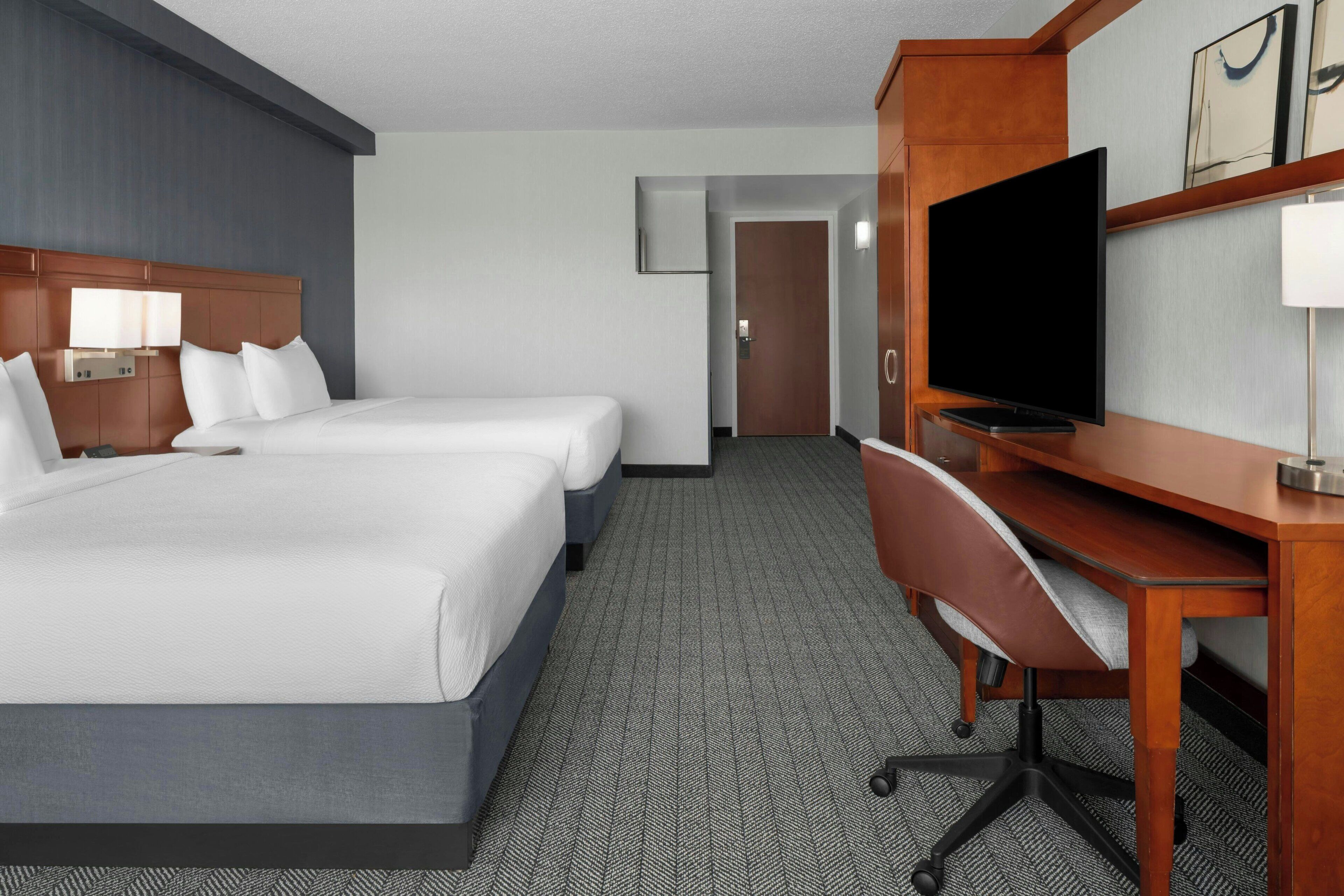 Quarto de hotel com duas camas, uma escrivaninha com cadeira, televisão e porta de acesso a outro espaço.