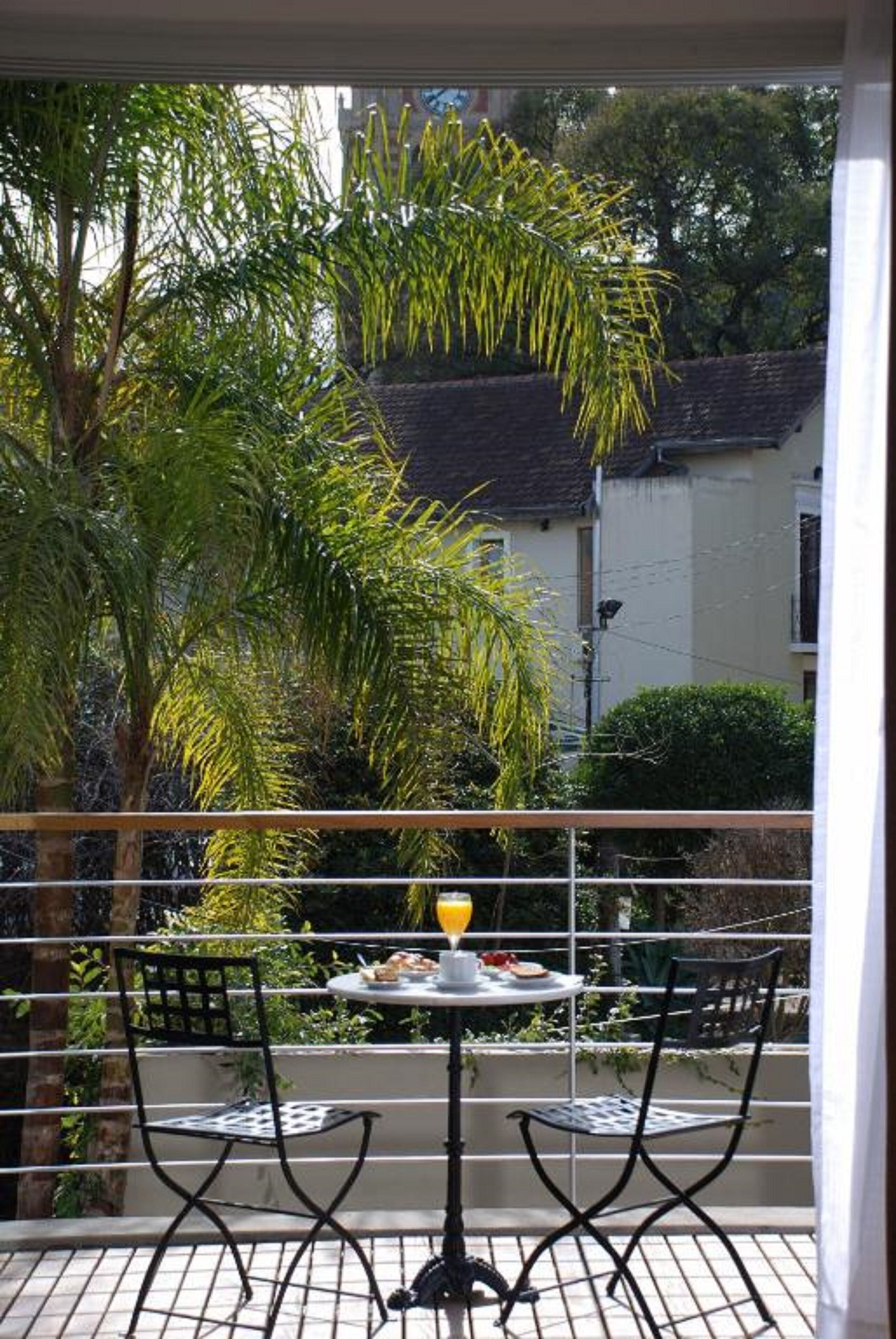 Uma varanda com mesa e cadeiras, dando vista para um jardim com palmeiras e um prédio ao fundo.