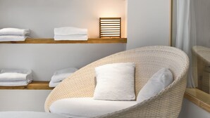 Um quarto com uma cadeira de vime, um banquinho redondo e prateleiras com toalhas dobradas.