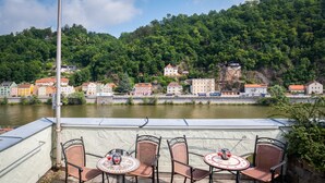 Um terraço com quatro cadeiras de vime e uma mesa pequena, com vista para um rio e edifícios na margem oposta.