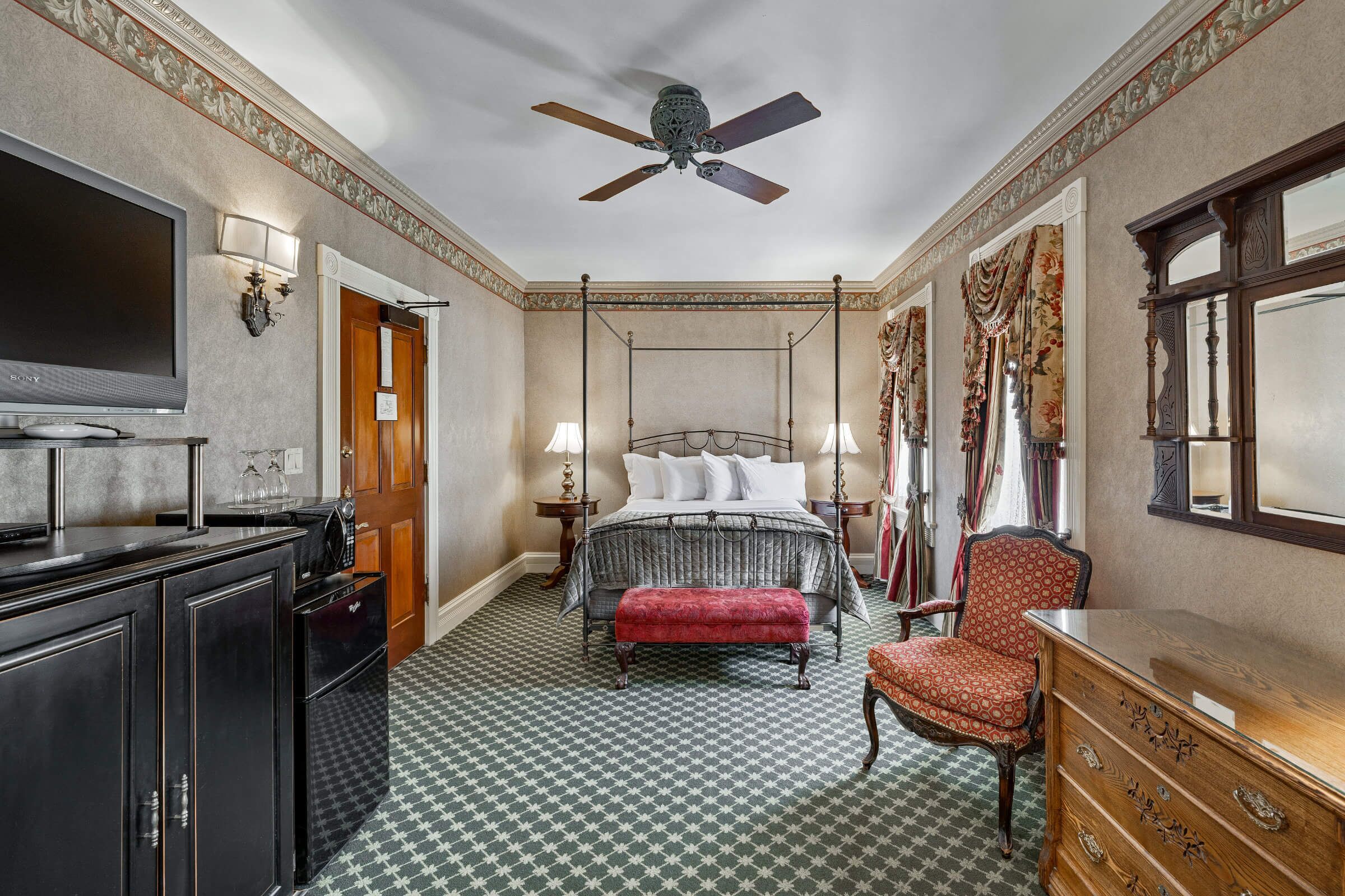 Quarto de hotel com uma cama de dossel, televisão, cómoda de madeira e uma cadeira estofada vermelha.
