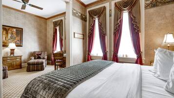 Quarto de hotel com uma cama grande, um ventilador de teto, uma escrivaninha com luminária, uma cadeira e um armário.