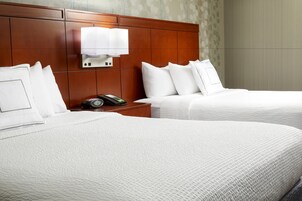 Quarto de hotel com duas camas, cabeceira de madeira, mesa de cabeceira com telefone e luminária fixada na parede.