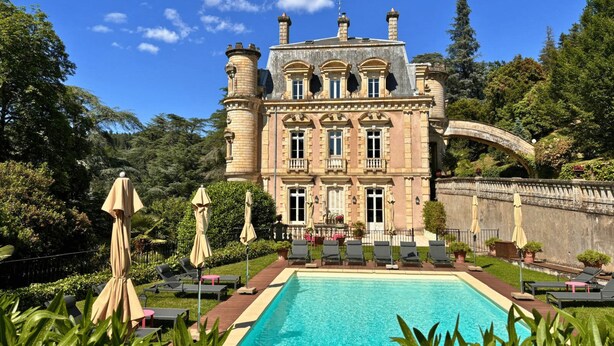 Um edifício grande e histórico com piscina, rodeado por vegetação exuberante e área de estar externa.
