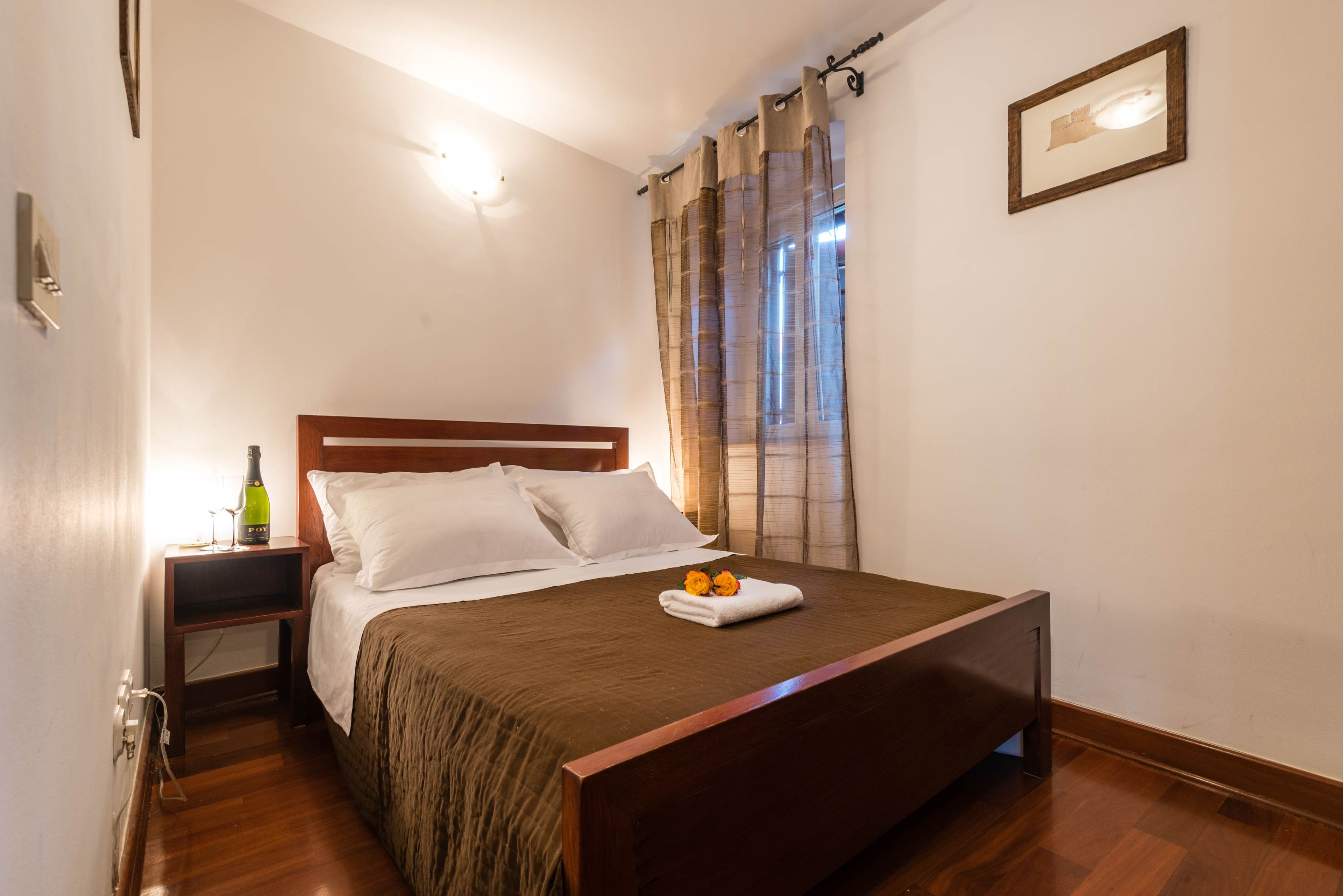 Quarto de hotel com cama de madeira, mesa de cabeceira, uma garrafa de champanhe e um buquê de flores.