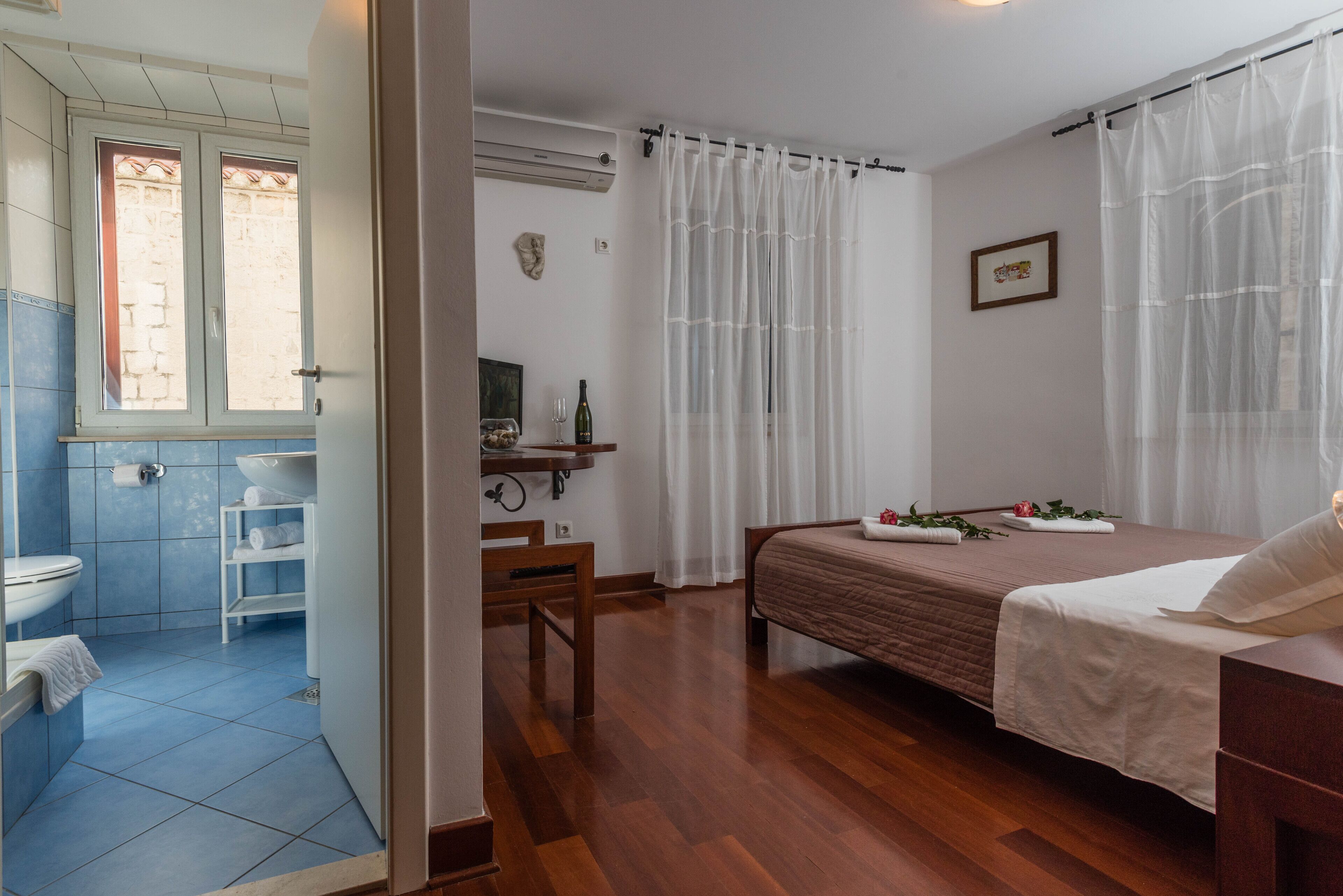Um quarto com piso de madeira, uma cama com colcha marrom, uma escrivaninha com cadeira e uma janela com cortina.