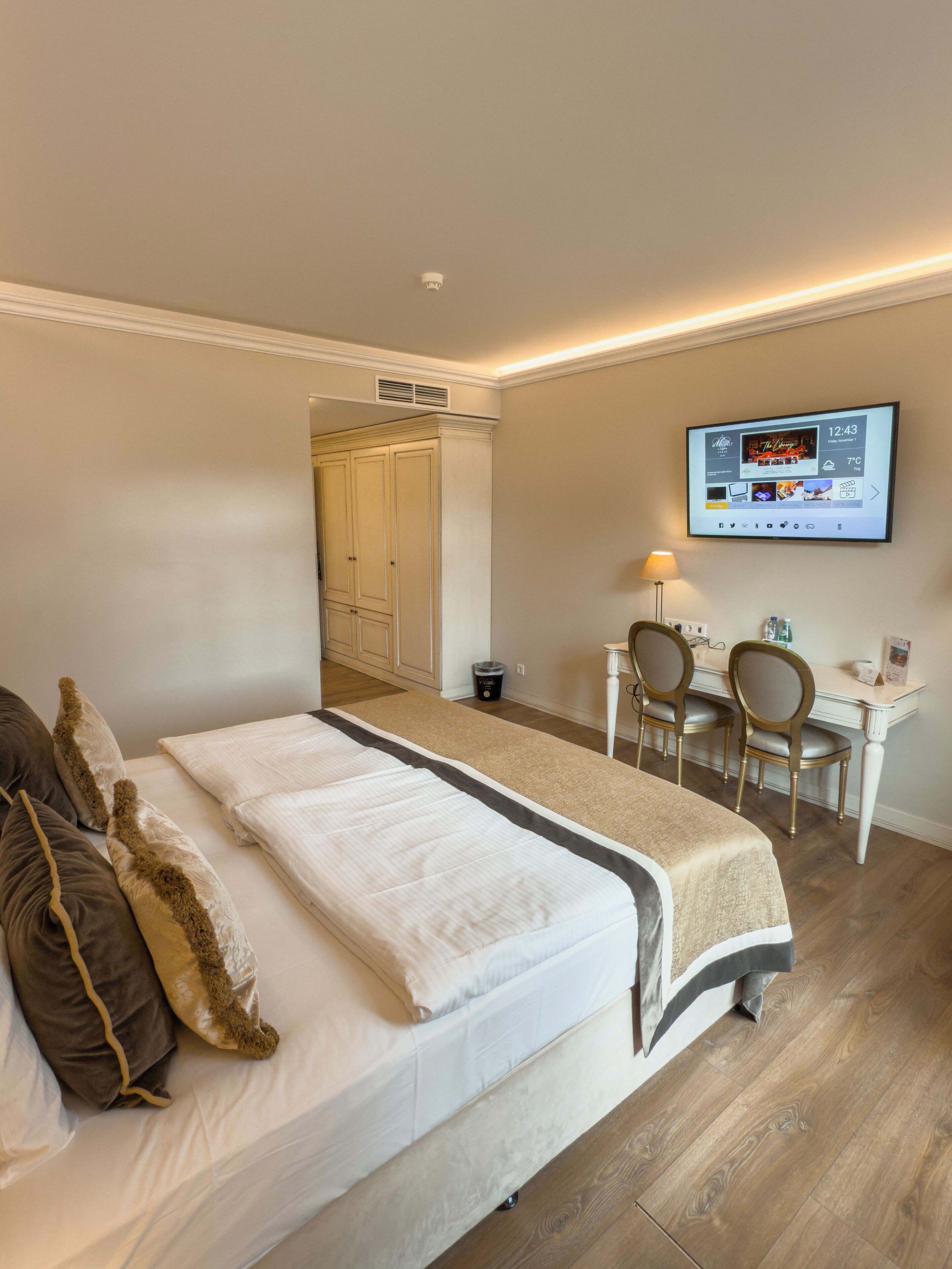 Quarto de hotel com cama, escrivaninha e televisão exibindo um programa.