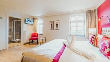Quarto de hotel com uma cama grande, uma cadeira vermelha, uma mesinha, televisão e área de chuveiro.