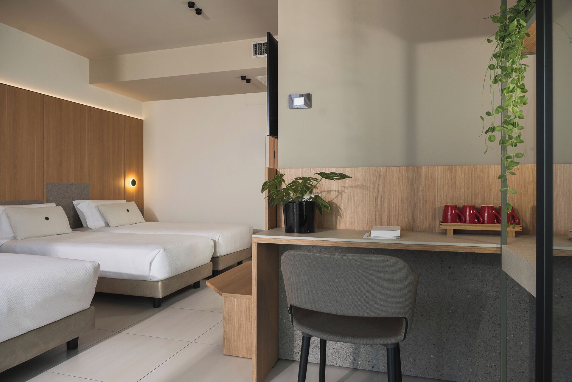Quarto de hotel moderno com duas camas, uma escrivaninha, uma cadeira, uma planta em vaso e uma prateleira com canecas vermelhas.