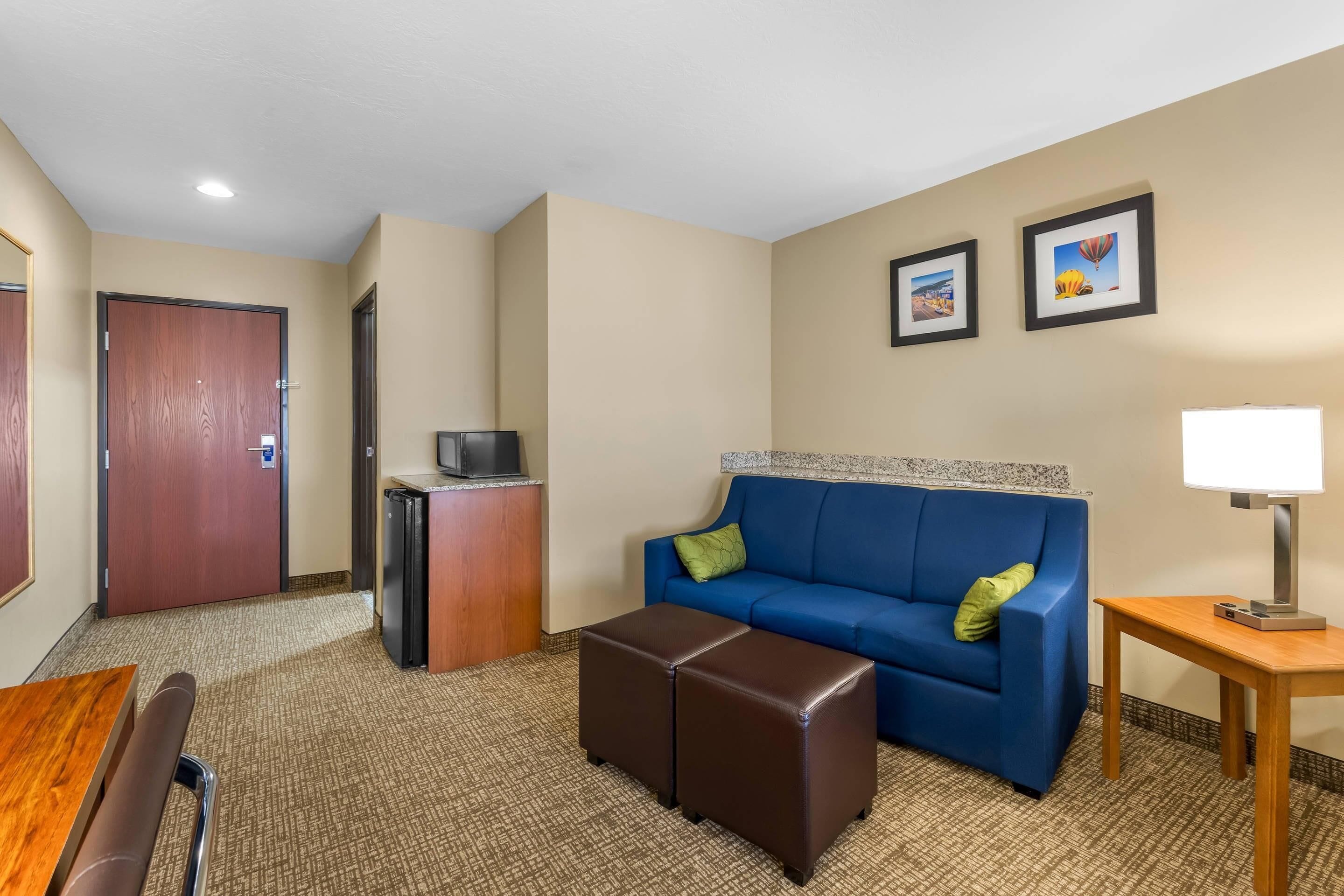 Quarto de hotel com sofá azul, mesa de madeira, pequena escrivaninha de madeira, abajur e porta.