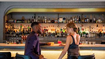 Uma área de bar com um homem e uma mulher sentados à mesa, um balcão de bar bem abastecido ao fundo e um design de teto curvo moderno.