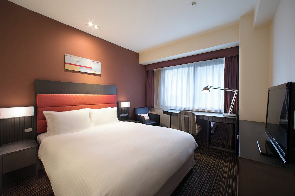 Quarto de hotel com uma cama grande, uma escrivaninha com cadeira, televisão e uma janela com cortinas.
