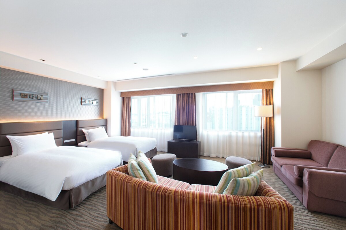 Quarto de hotel com duas camas, um sofá, uma mesa de centro redonda, uma televisão e uma janela grande com cortinas.