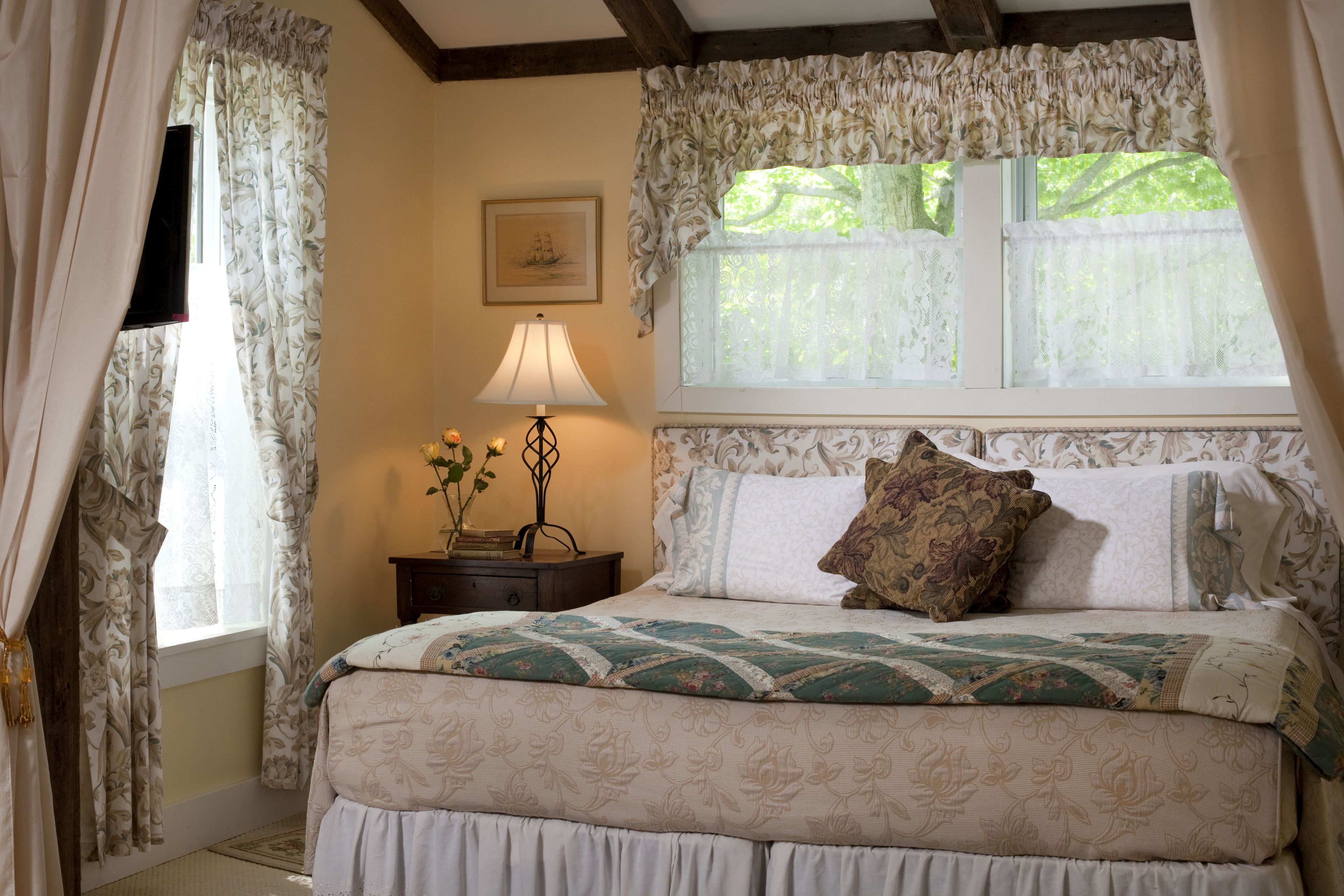 Quarto com uma cama grande, cortinas florais, mesa de cabeceira com abajur e janela com vista para o verde.