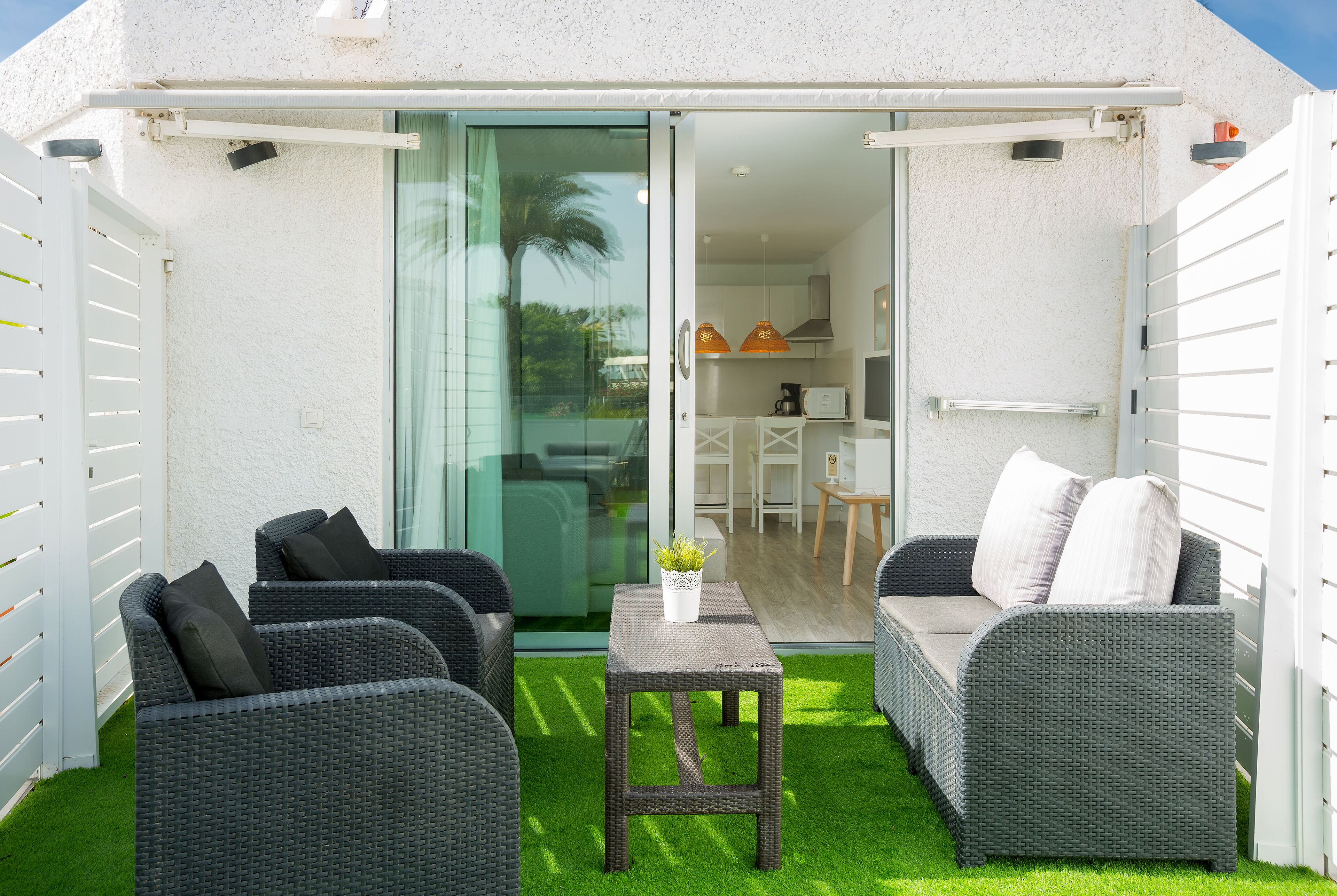 Um pátio moderno ao ar livre com móveis de vime, uma pequena mesa com uma planta e uma porta de vidro deslizante que dá para um espaço interno.