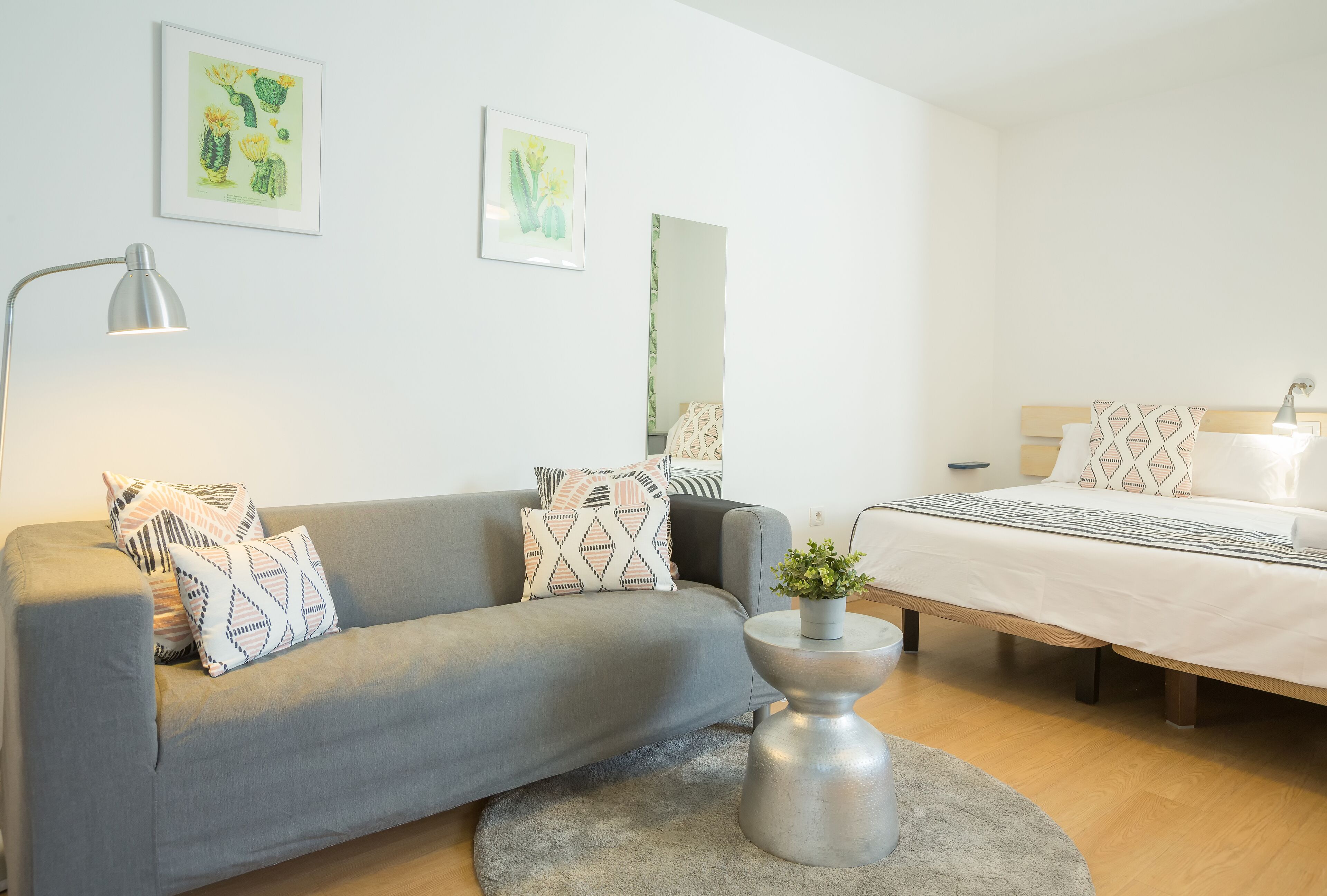 Um quarto moderno com um sofá cinzento, uma mesa de cabeceira com uma planta, uma cama com roupa de cama estampada e duas impressões botânicas emolduradas na parede.