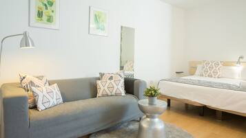Um quarto moderno com um sofá cinzento, uma mesa de cabeceira com uma planta, uma cama com roupa de cama estampada e duas impressões botânicas emolduradas na parede.