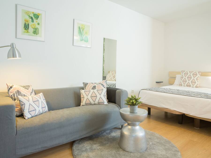 Um quarto moderno com um sofá cinzento, uma mesa de cabeceira com uma planta, uma cama com roupa de cama estampada e duas impressões botânicas emolduradas na parede.