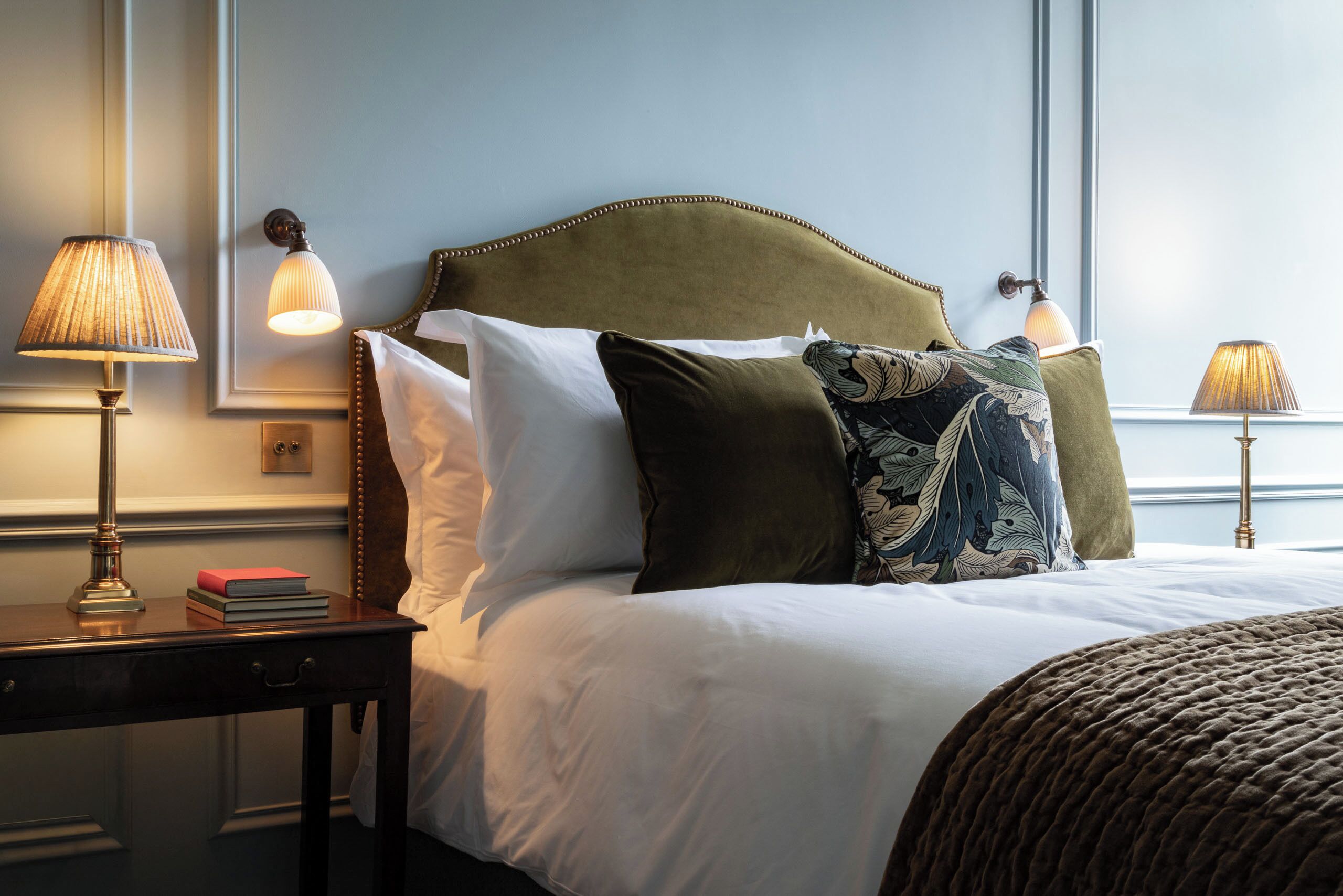 Cama bem arrumada com cabeceira verde, roupa de cama branca e almofadas decorativas. Existem duas luminárias de cabeceira de cada lado da cama.