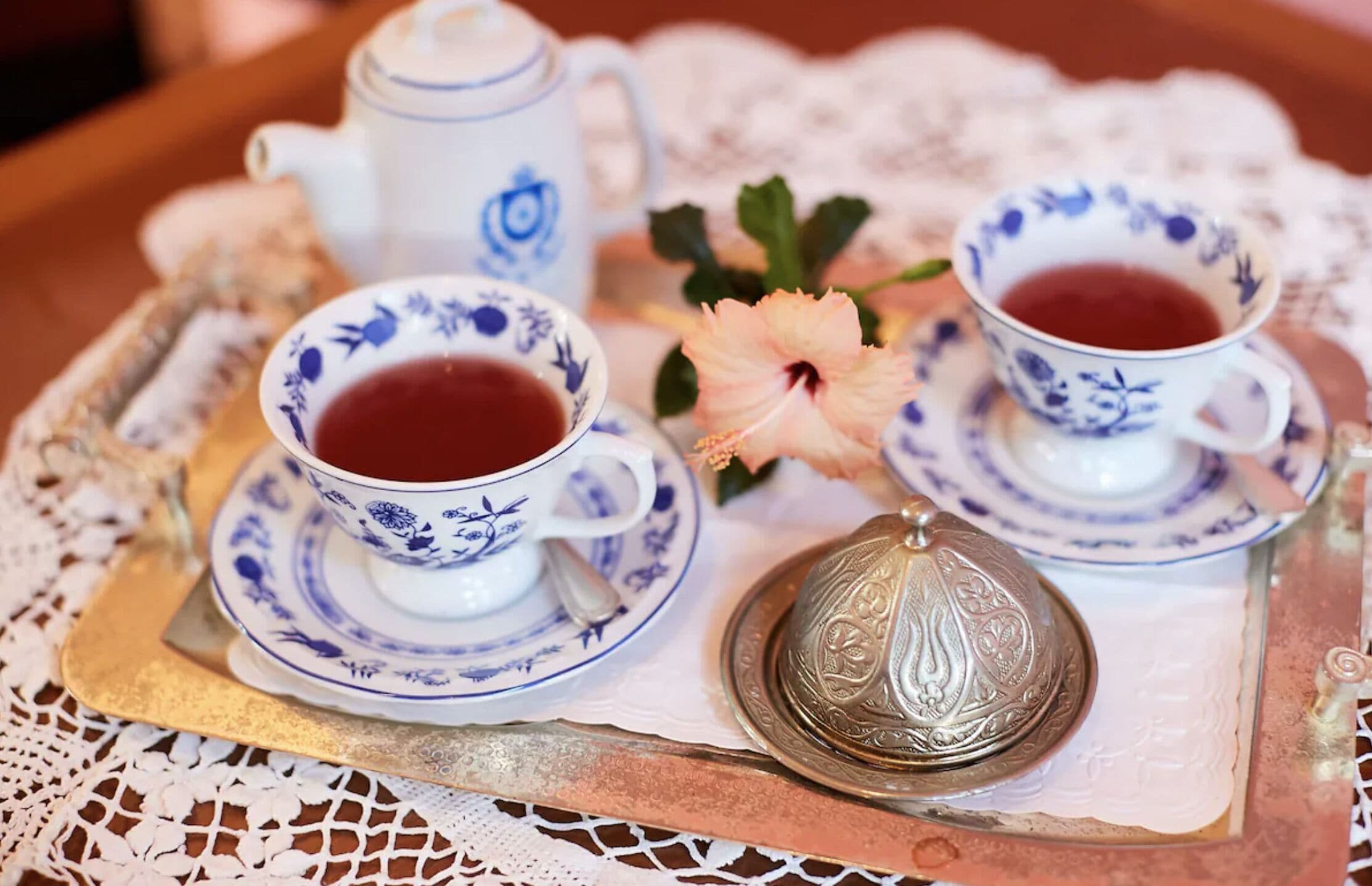 Uma bandeja com duas xícaras de chá, um bule e um objeto metálico decorativo sobre uma toalha de renda.