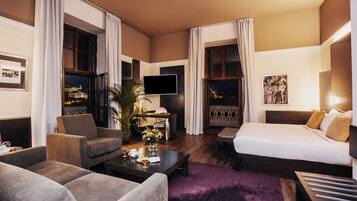 Quarto de hotel com sofá, cama, televisão, mesa de jantar e janela com cortinas.