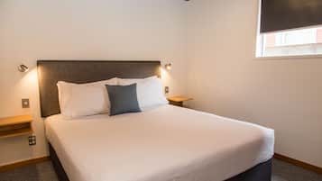 Uma cama bem arrumada com cabeceira escura e roupa de cama branca, duas mesas de cabeceira com luminárias e uma janela com cortina.