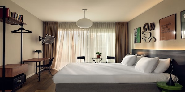Quarto de hotel com uma cama grande, uma escrivaninha com cadeira, estante de livros, televisão e janela com cortinas.