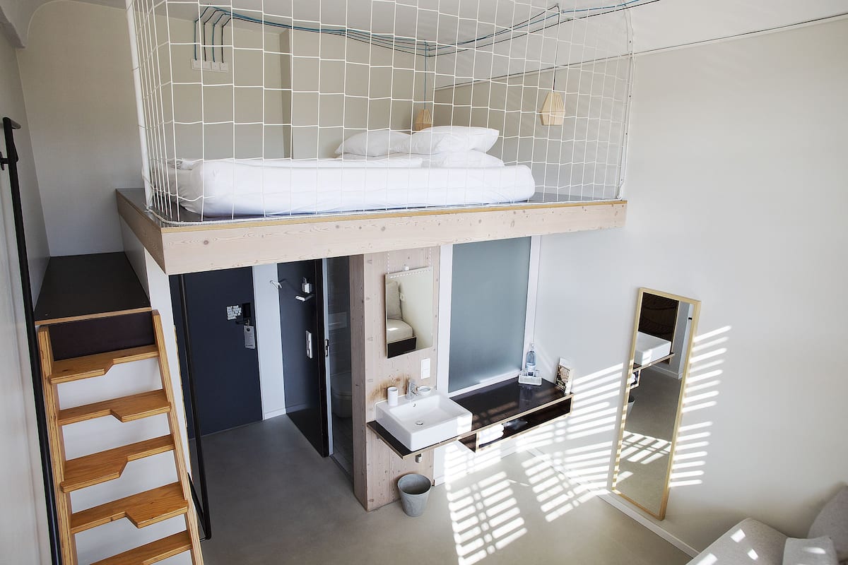 Uma cama beliche com colchão branco, escada de madeira, pia e espelho.