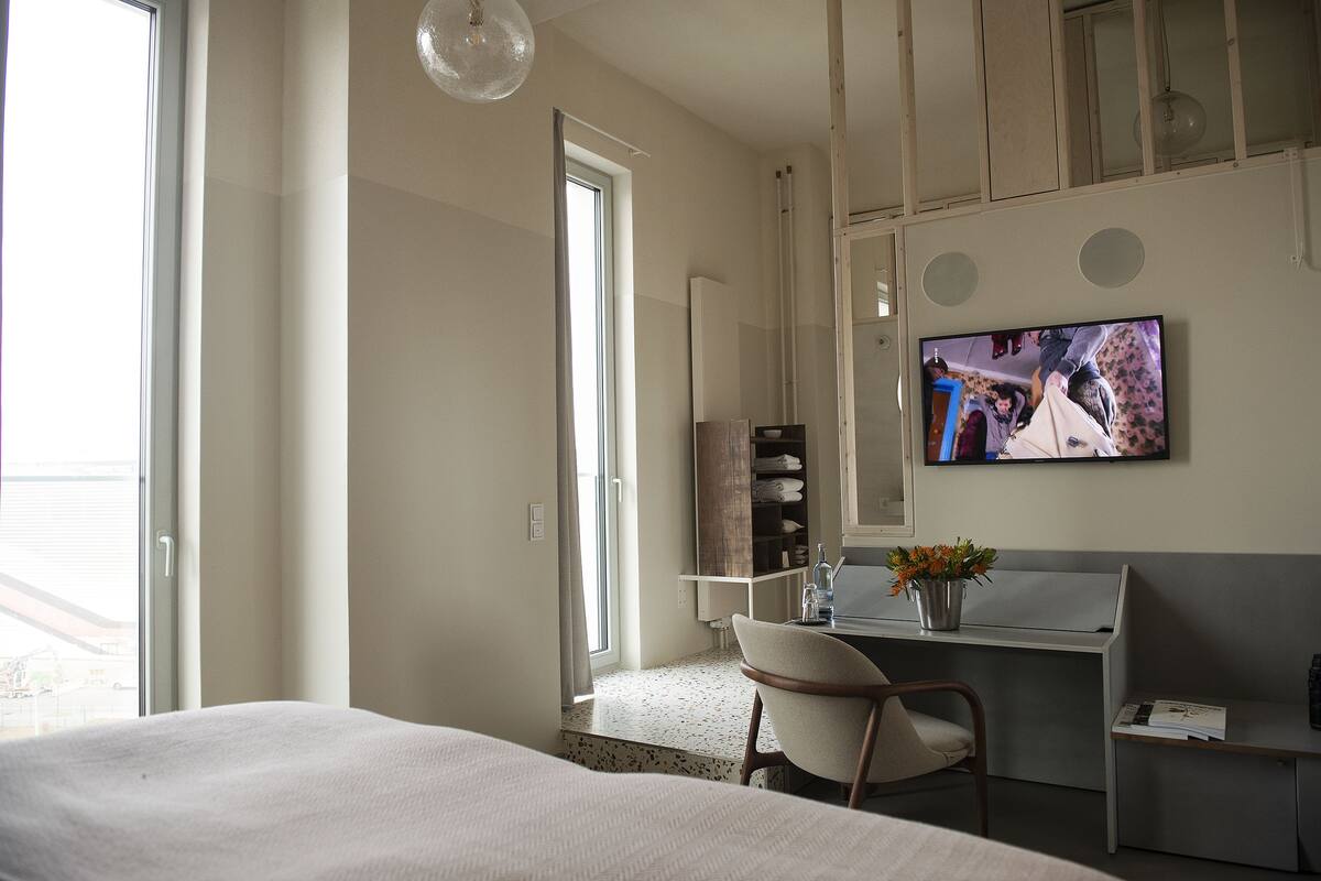 Quarto de hotel com cama, escrivaninha, cadeira, televisão e janela.