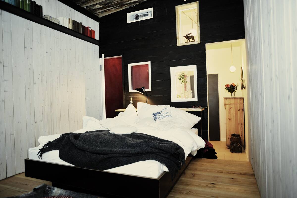 Quarto com cama, piso de madeira, parede branca, porta, espelho e prateleira com livros.