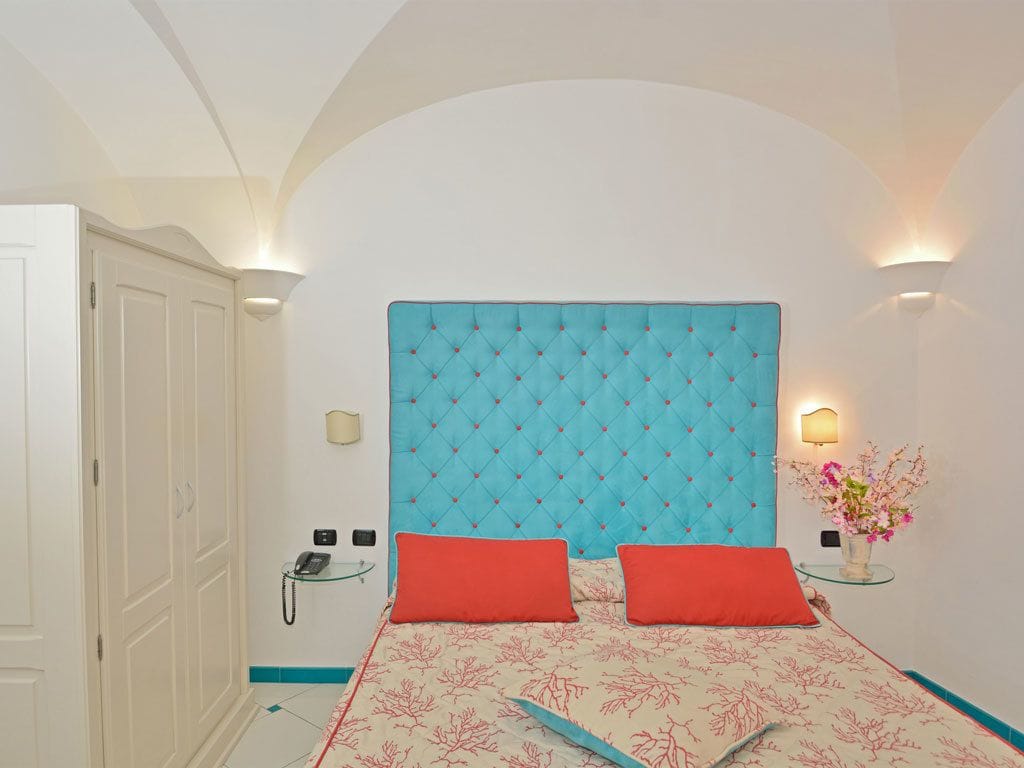 Quarto com cabeceira estofada, duas almofadas vermelhas, mesa de cabeceira com abajur e um vaso de flores.