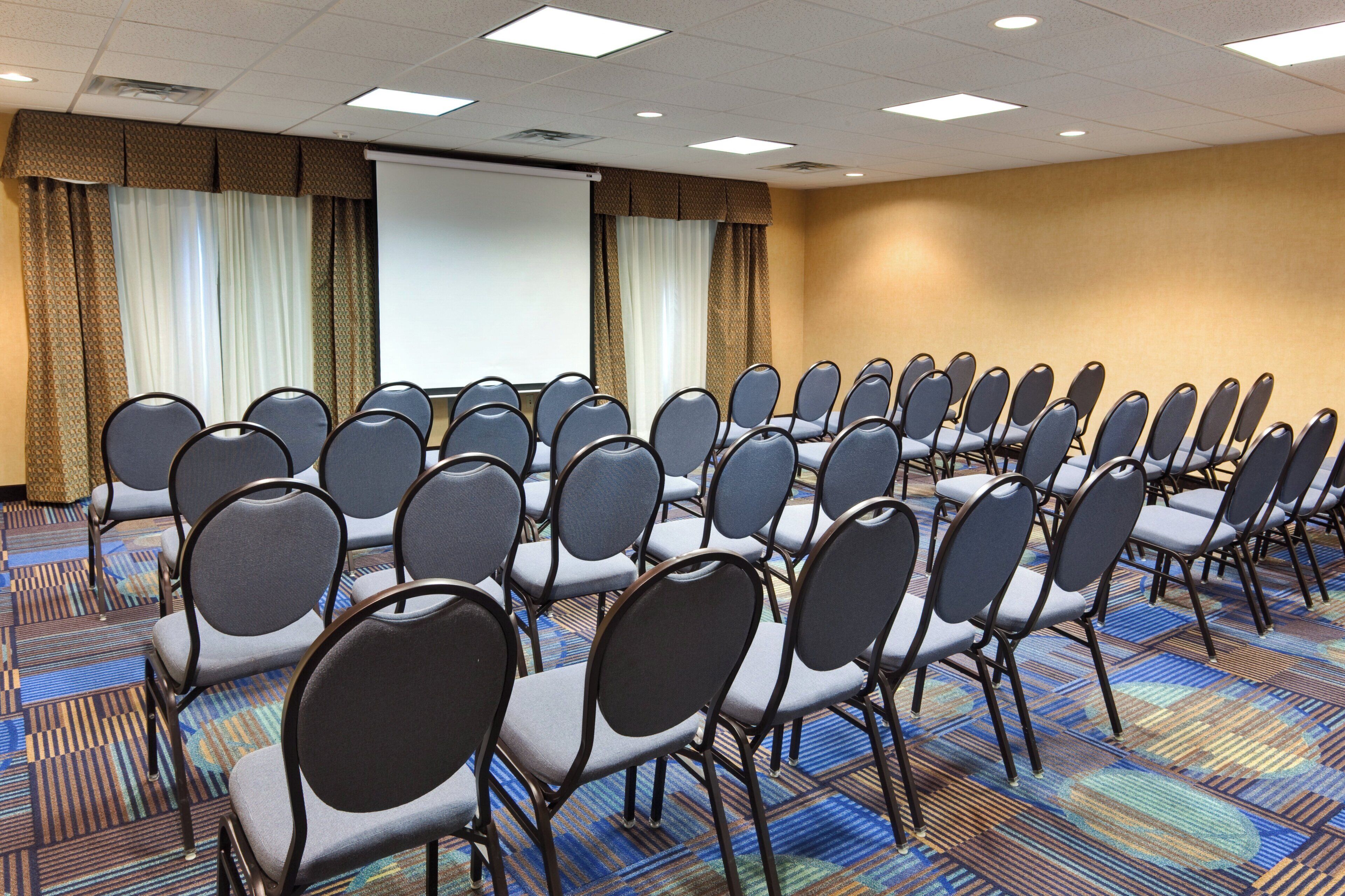Sala de conferência com fileiras de cadeiras voltadas para uma tela de projeção, piso coberto de carpete e cortinas nas janelas.