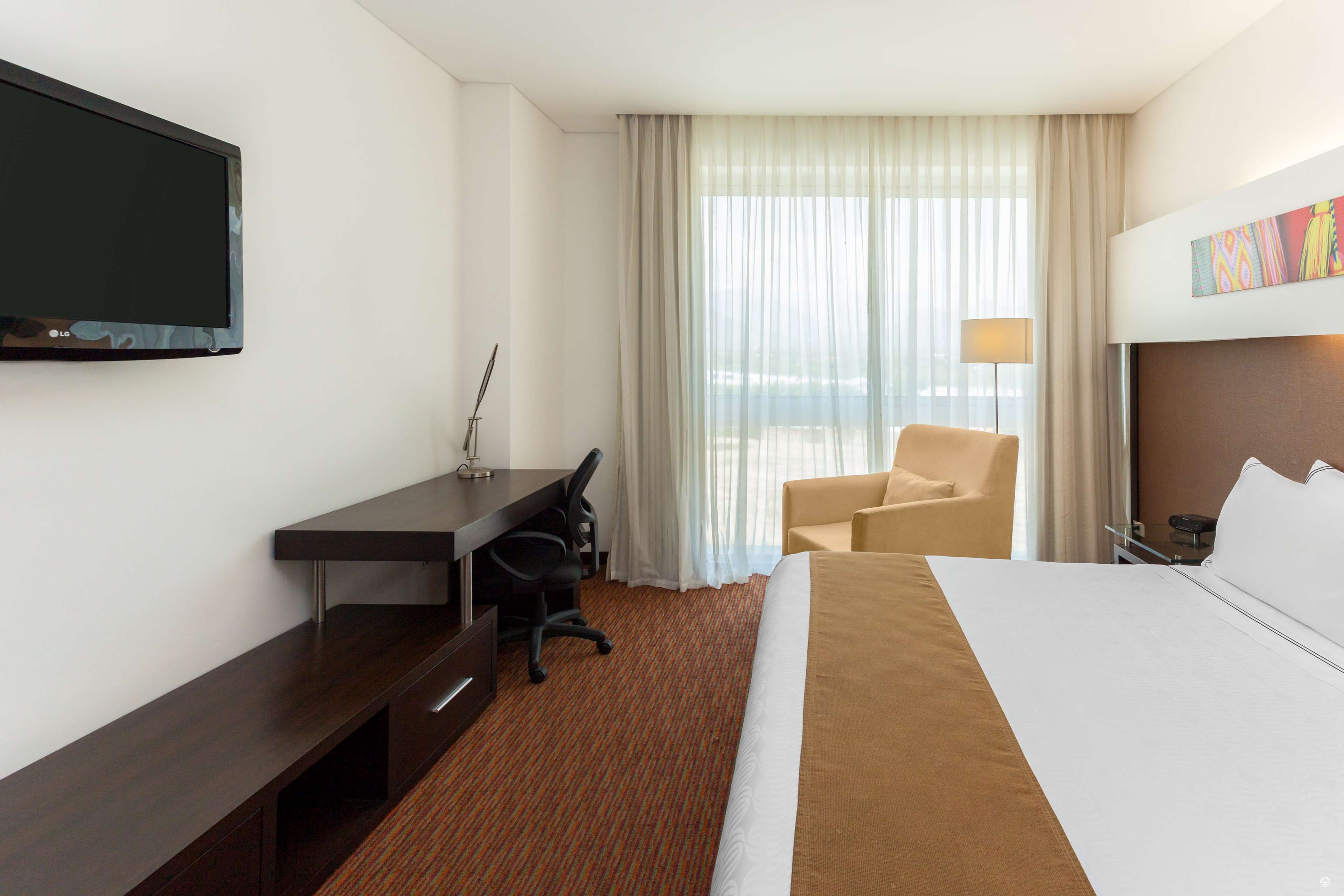 Quarto de hotel com cama, escrivaninha, cadeira, televisão e janela com cortinas.