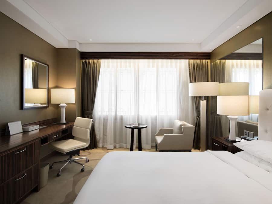 Quarto de hotel com uma cama grande, duas cadeiras, uma escrivaninha com computador, televisão e janela com cortinas.