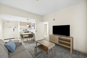 Quarto de hotel com sofá, mesa de centro, televisão em suporte, cozinha compacta e área de refeições.