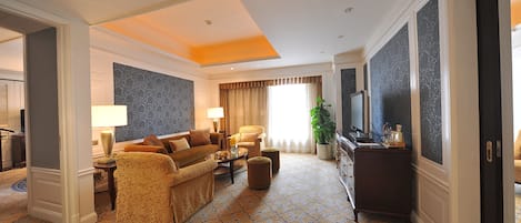 Quarto de hotel com área de estar, televisão, escrivaninha e uma grande janela com cortinas.