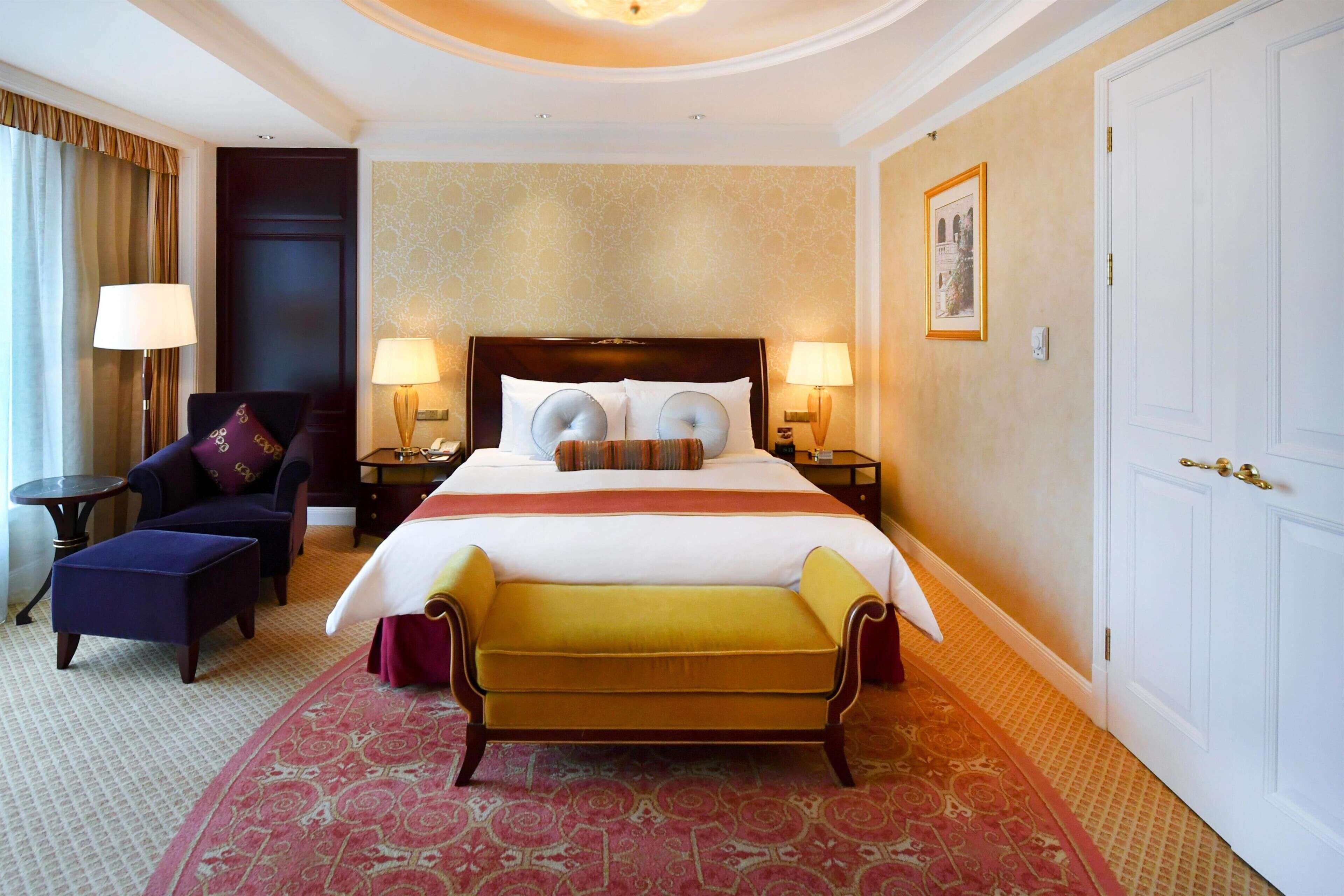 Quarto de hotel com uma cama grande, duas mesas de cabeceira com luminárias, um sofá, um banco e uma porta.